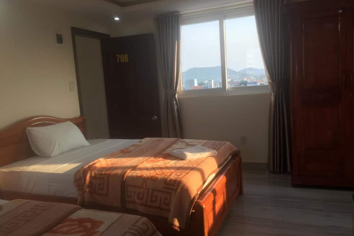 Quarto de hotel com duas camas, uma janela grande com cortinas, uma porta com número e vista para a cidade.