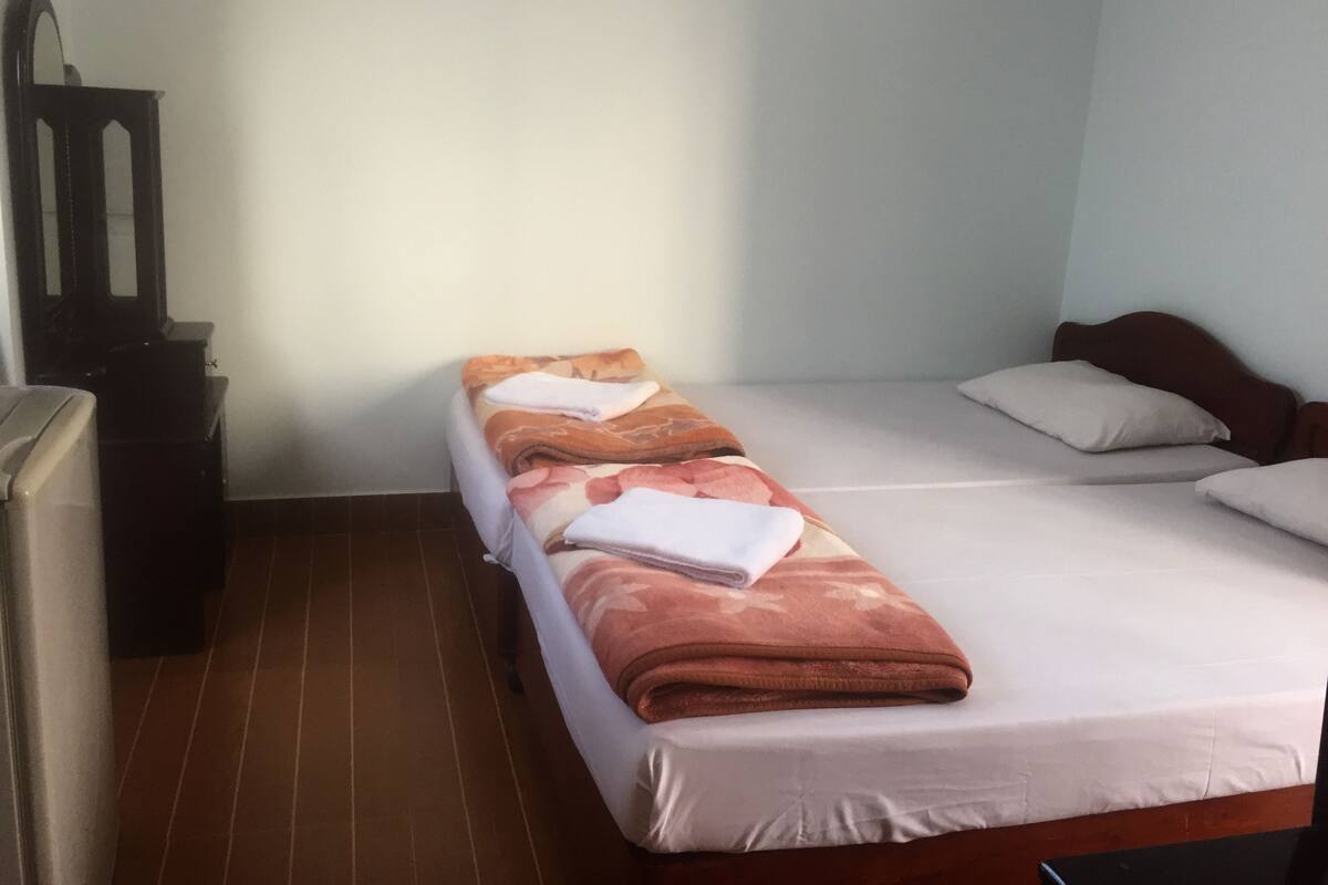 Uma cama de solteiro com colchão branco e cobertor marrom, cabeceira de madeira e uma pequena mesa de cabeceira com espelho.