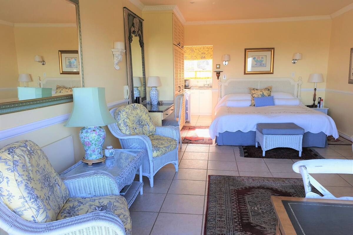 Quarto de hotel com cama, duas cadeiras de vime branco, uma mesa, um espelho grande e uma mesinha de cabeceira com abajur.