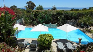 Uma área de piscina com espreguiçadeiras e guarda-sóis, rodeada por vegetação exuberante e vista para o mar.