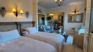 Quarto de hotel com duas camas, área de estar com mesa e cadeiras, cozinha ao fundo e luminárias decorativas na parede.