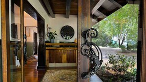 Uma área de recepção com um banco de madeira, um relógio de parede grande e vista para um jardim através de uma porta aberta.