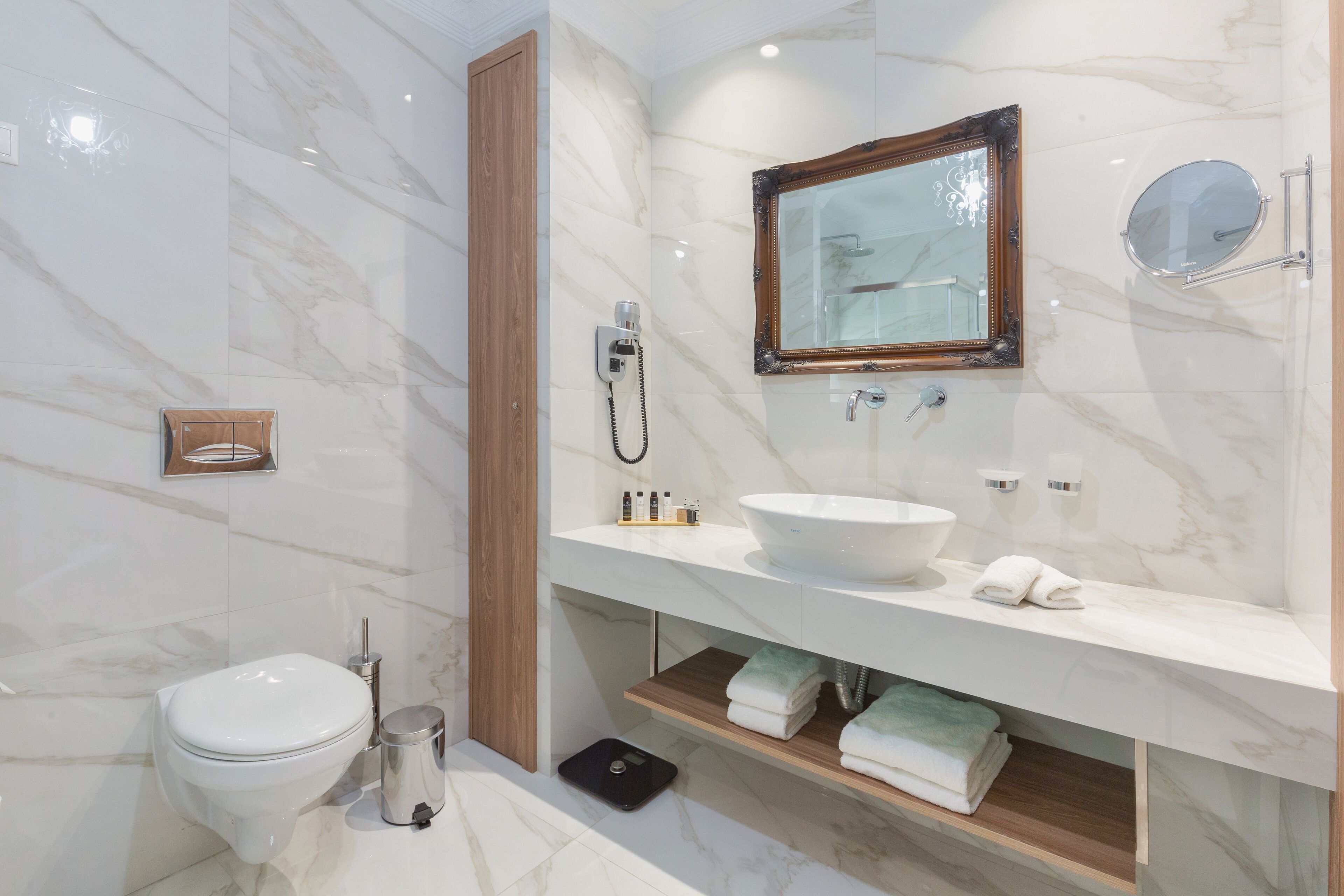 Um banheiro moderno com vaso sanitário branco, pia com bancada de mármore, aparador de madeira, espelho e porta-toalhas.