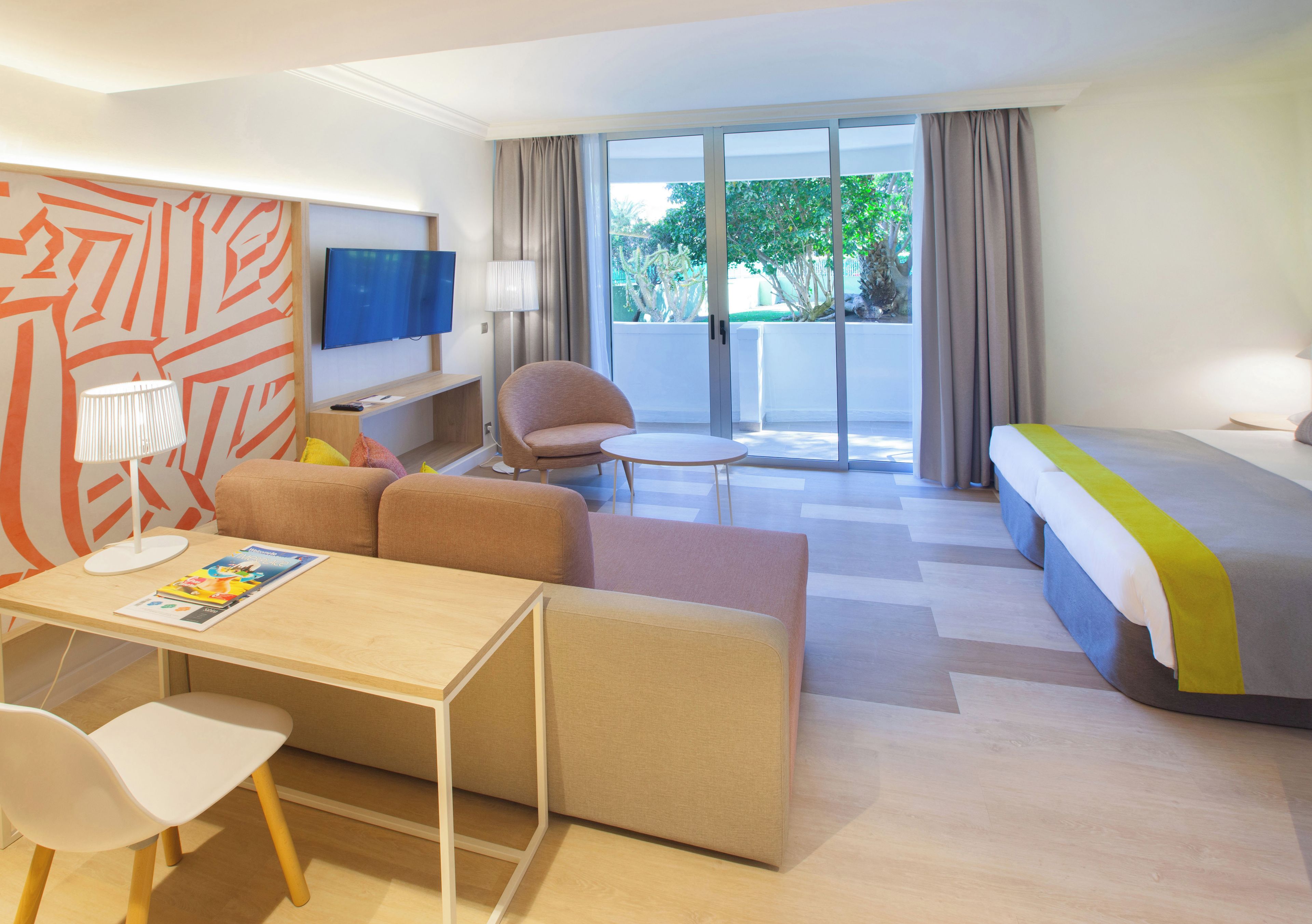 Quarto de hotel com sofá, mesa pequena, televisão, cama e varanda com vista para área verde.
