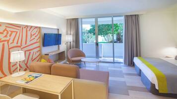 Quarto de hotel com sofá, mesa pequena, televisão, cama e varanda com vista para área verde.