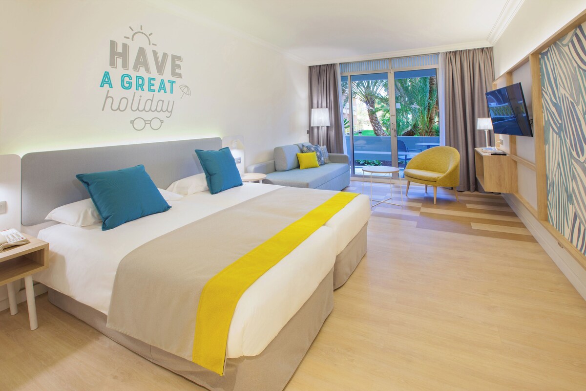 Quarto de hotel com cama grande, sofá, poltrona, televisão e varanda com cortinas.