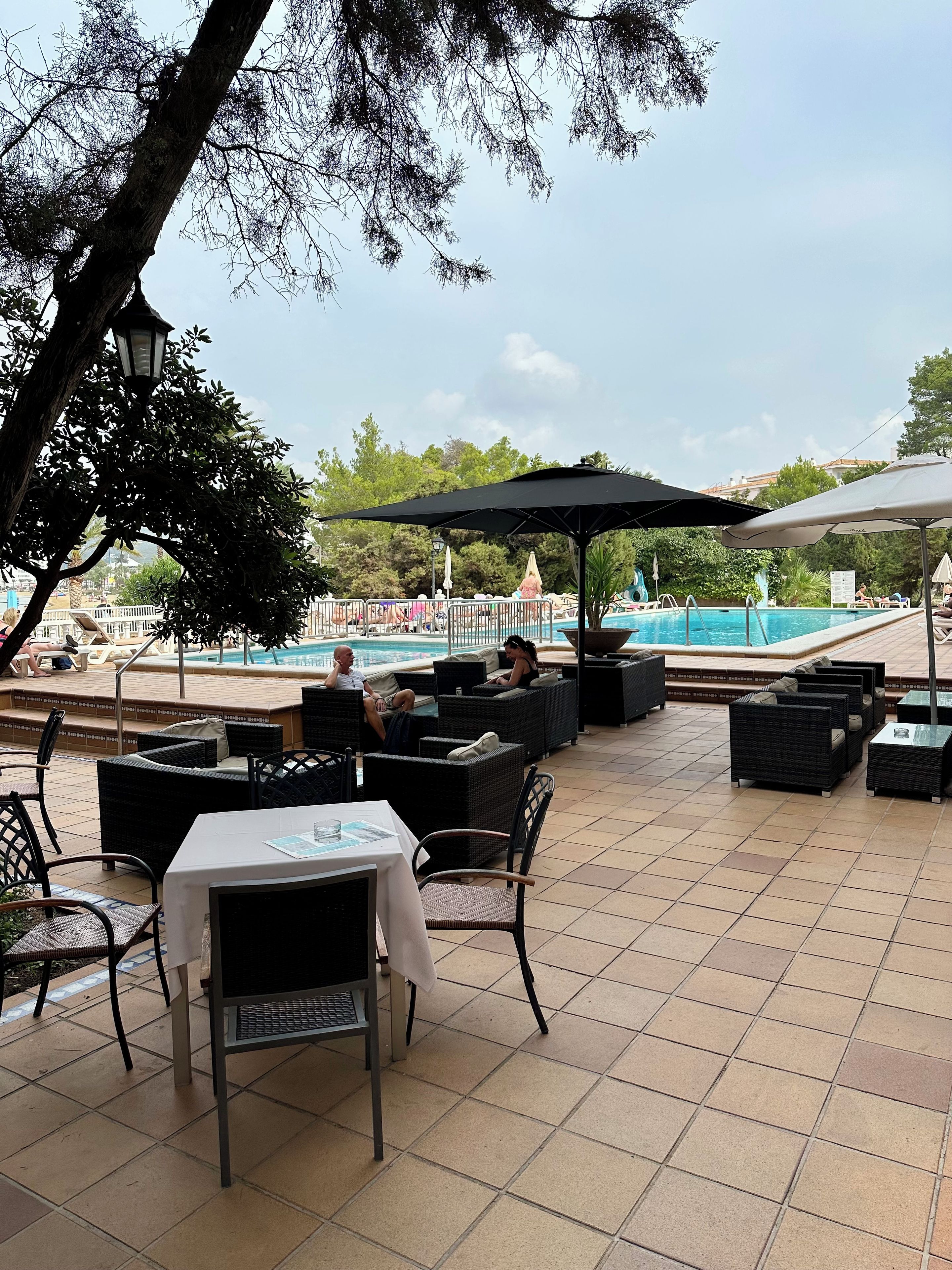 Um pátio externo com assentos e mesas, piscina ao fundo e vista da região.