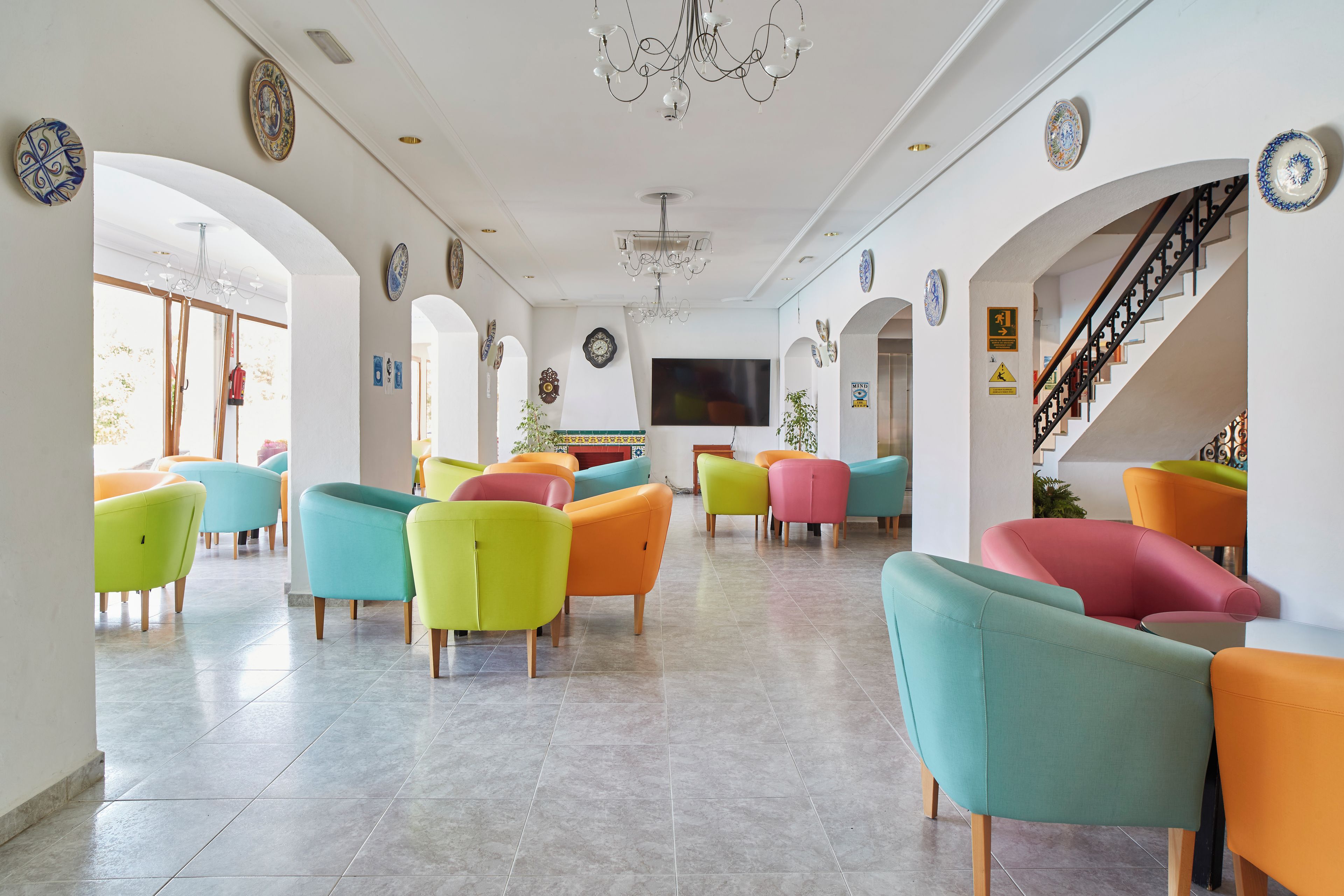 Um hall amplo com poltronas coloridas, um lustre e uma escadaria.
