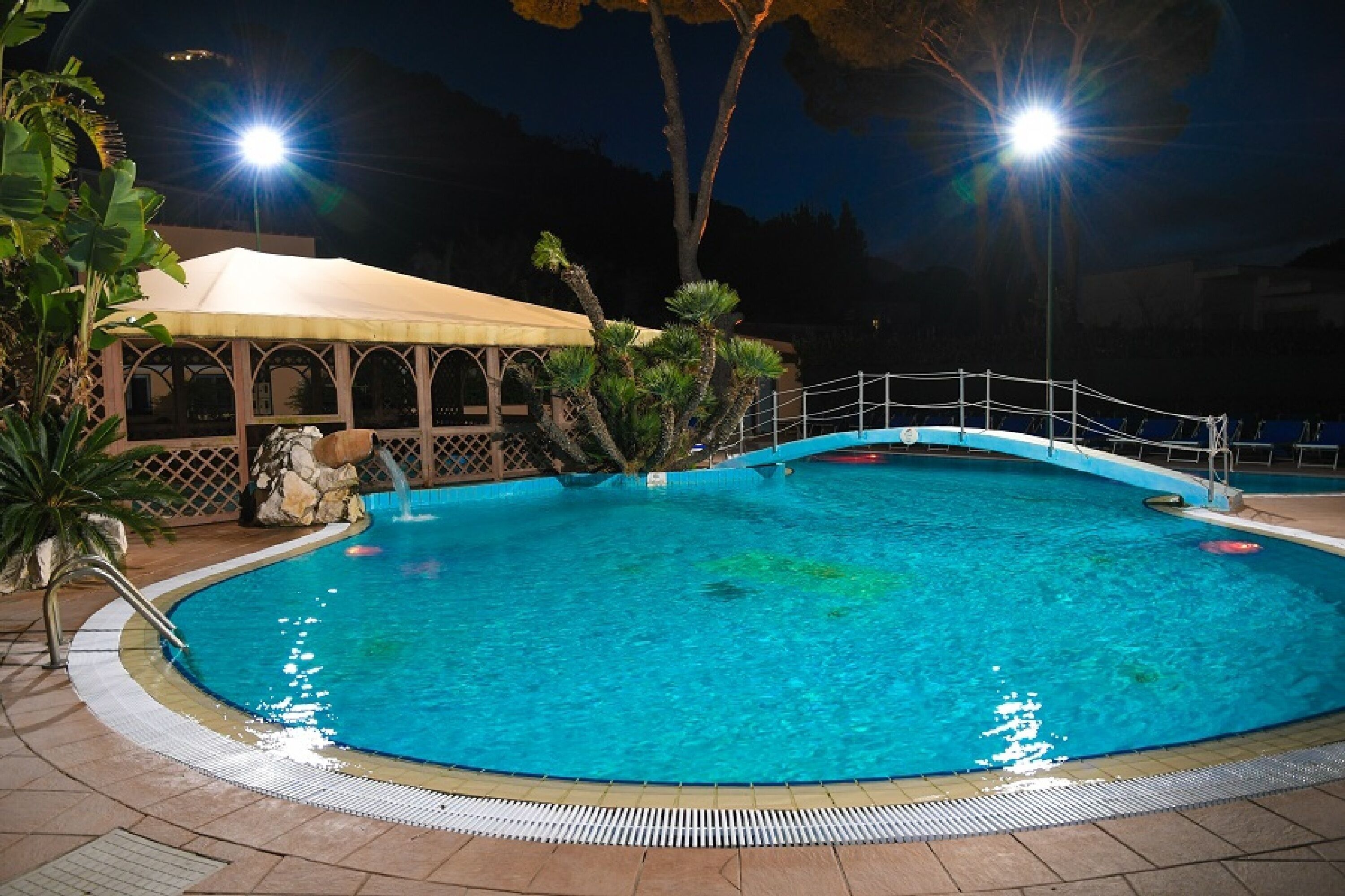 Uma piscina com cerca branca, área de estar coberta e palmeiras.
