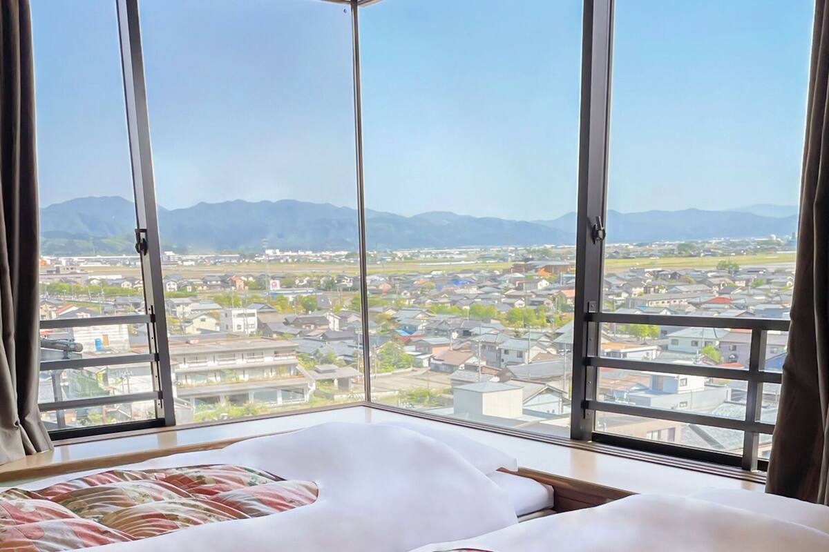 Quarto de hotel com uma janela ampla, uma cama e vista para uma cidade e montanhas.