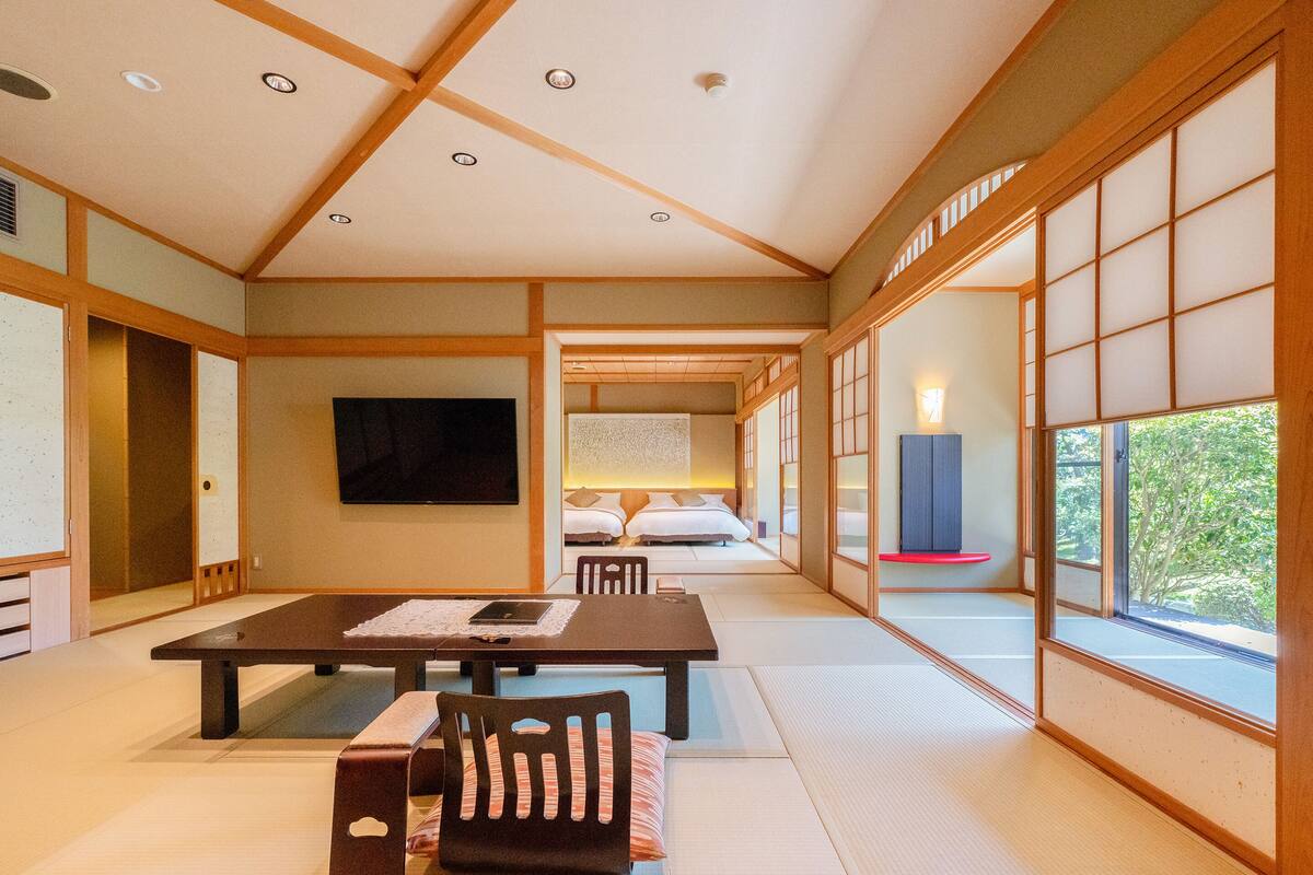 Um quarto em estilo tradicional japonês, com piso de tatame, uma mesa baixa com almofada, televisão na parede e vista para um jardim através de grandes portas deslizantes.
