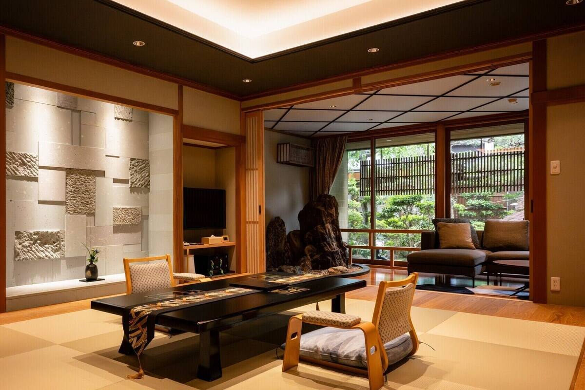 Um quarto japonês tradicional com piso de tatame, uma mesa baixa com um jogo de mesa preto, uma televisão e vista para um jardim através de grandes janelas.