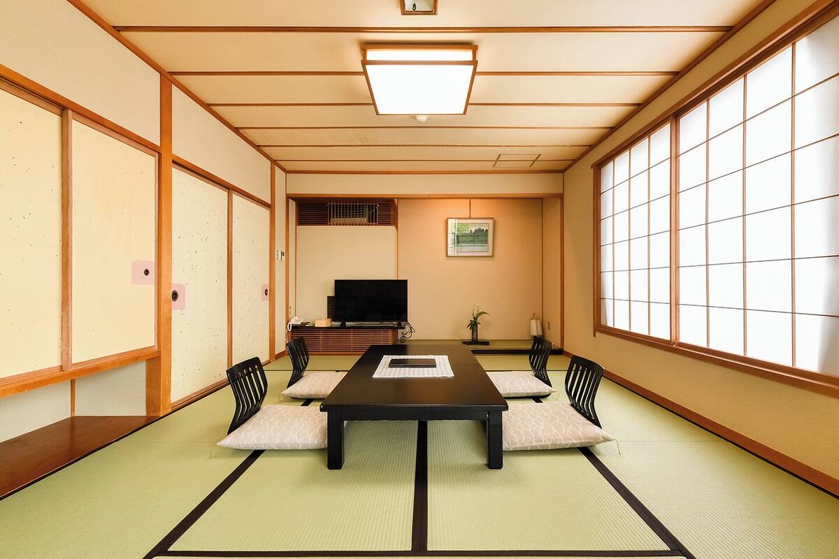 Quarto japonês tradicional com tatames, uma mesa baixa, cadeiras, televisão e uma janela grande.