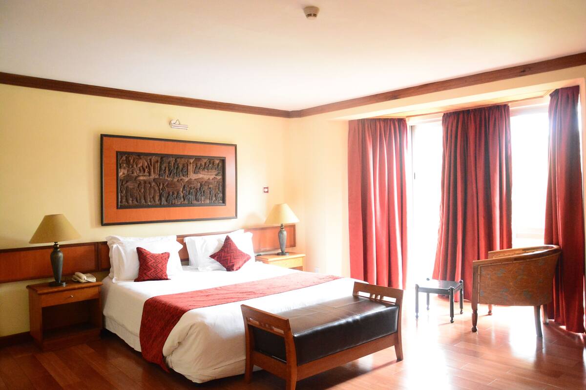Quarto de hotel com uma cama grande, duas mesas de cabeceira com luminárias, um banco, uma cadeira e um quadro grande na parede.