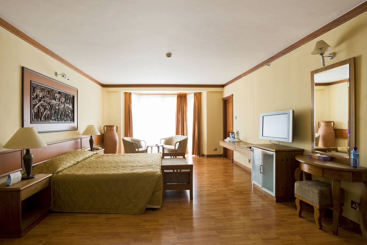 Quarto de hotel com cama, escrivaninha, cadeira, televisão e uma janela grande com cortinas.