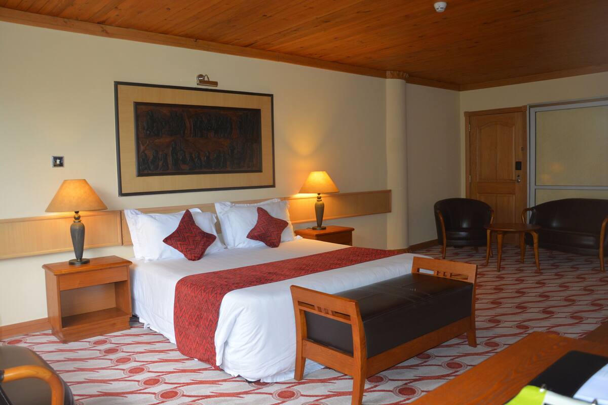 Quarto de hotel com cama, duas mesas de cabeceira com luminárias, um banco de madeira, uma cadeira e um quadro grande na parede.
