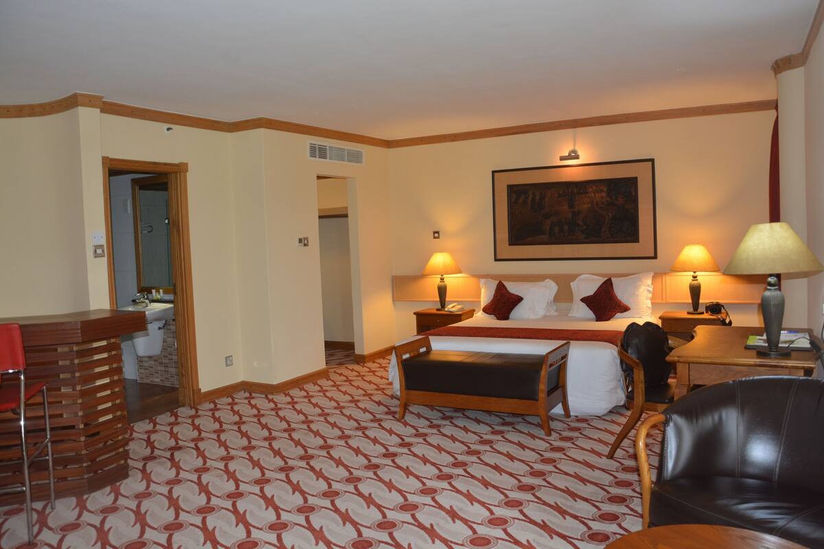 Quarto de hotel com uma cama, duas luminárias de cabeceira, uma escrivaninha com cadeira, uma mesinha e um tapete estampado.