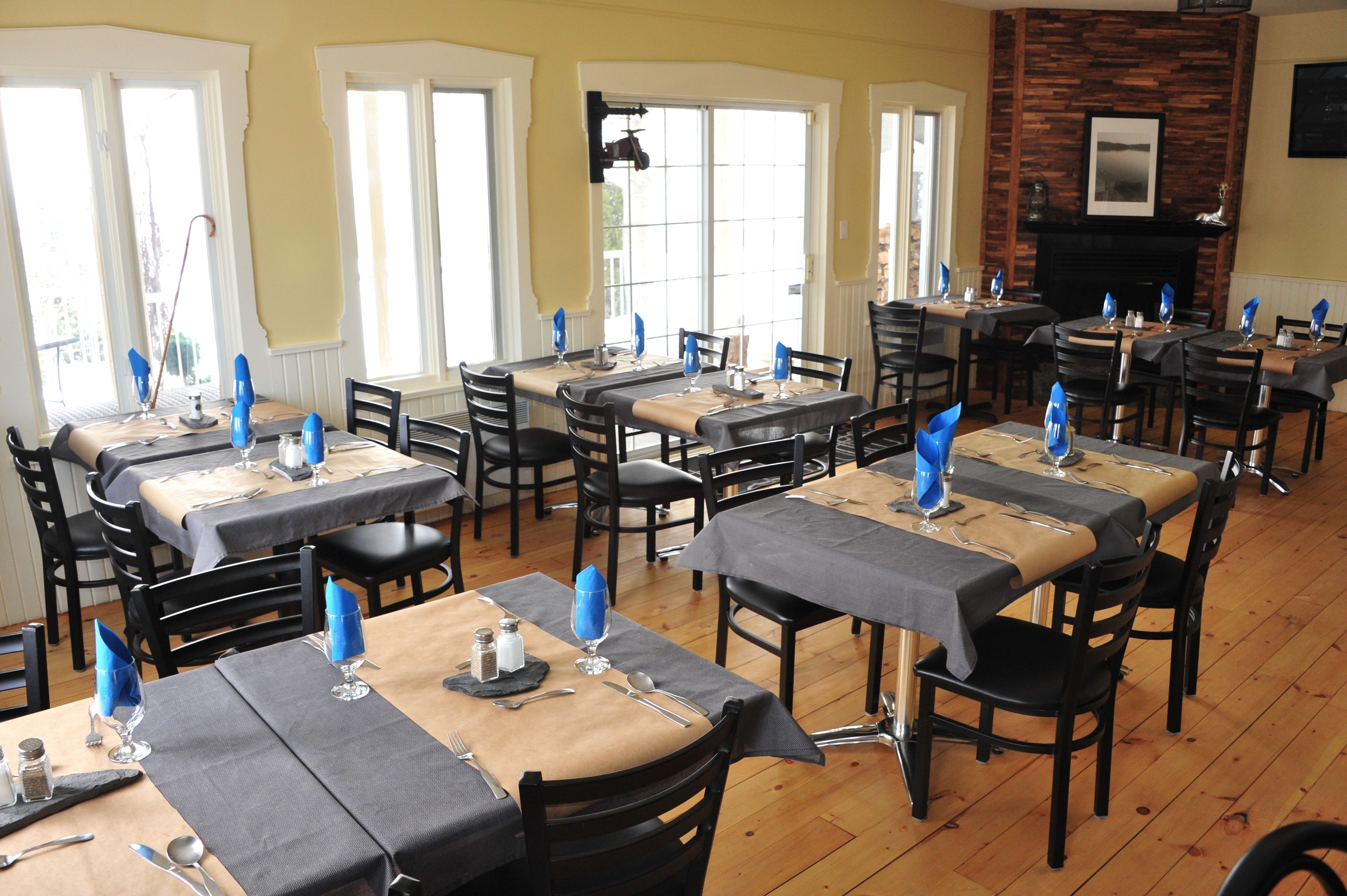 Uma sala de jantar com várias mesas preparadas para o serviço, com cadeiras pretas e toalhas de mesa bege.