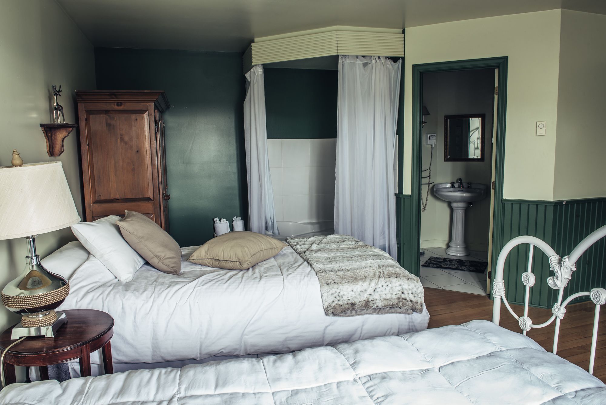 Quarto com cama, mesa de cabeceira com abajur, guarda-roupa, banheiro com pia e espelho, e chuveiro.