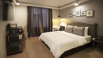 Quarto de hotel com uma cama grande, uma escrivaninha com cadeira, uma televisão e uma janela com cortinas.