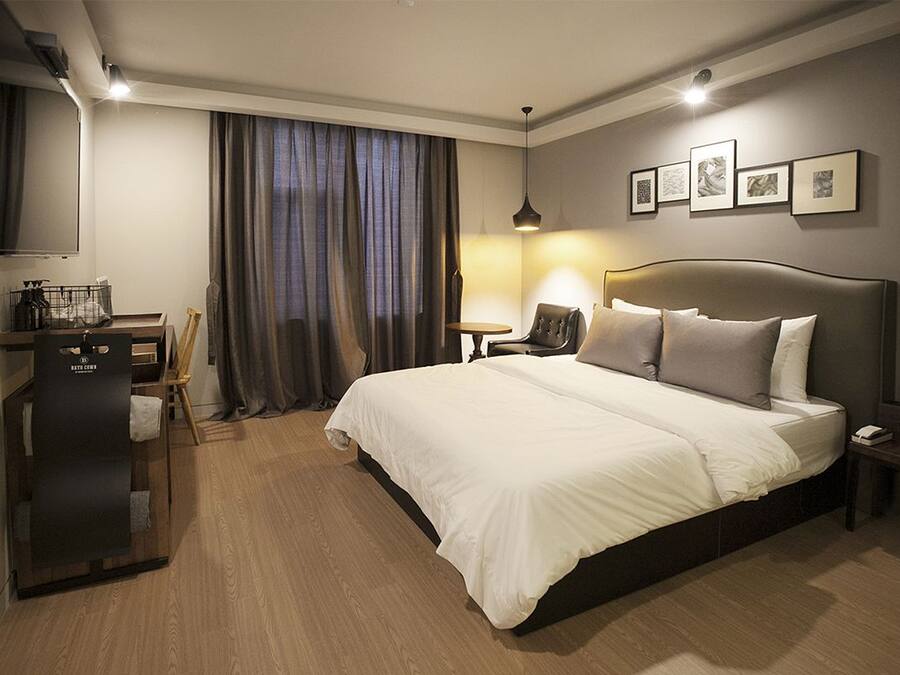 Quarto de hotel com uma cama grande, uma escrivaninha com cadeira, uma televisão e uma janela com cortinas.