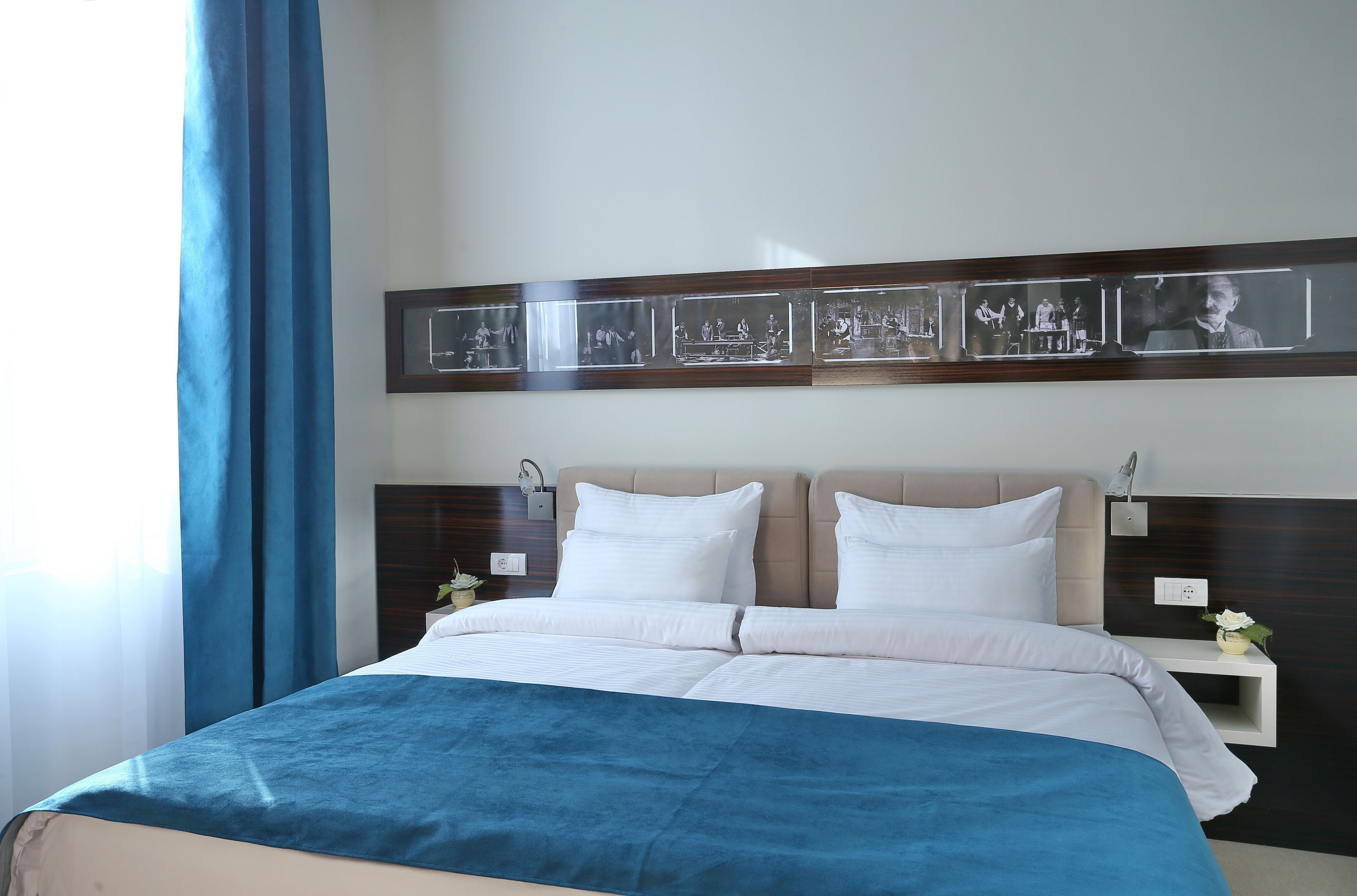 Quarto de hotel com cama, mesa de cabeceira, painel de fotos na parede e janela com cortinas.