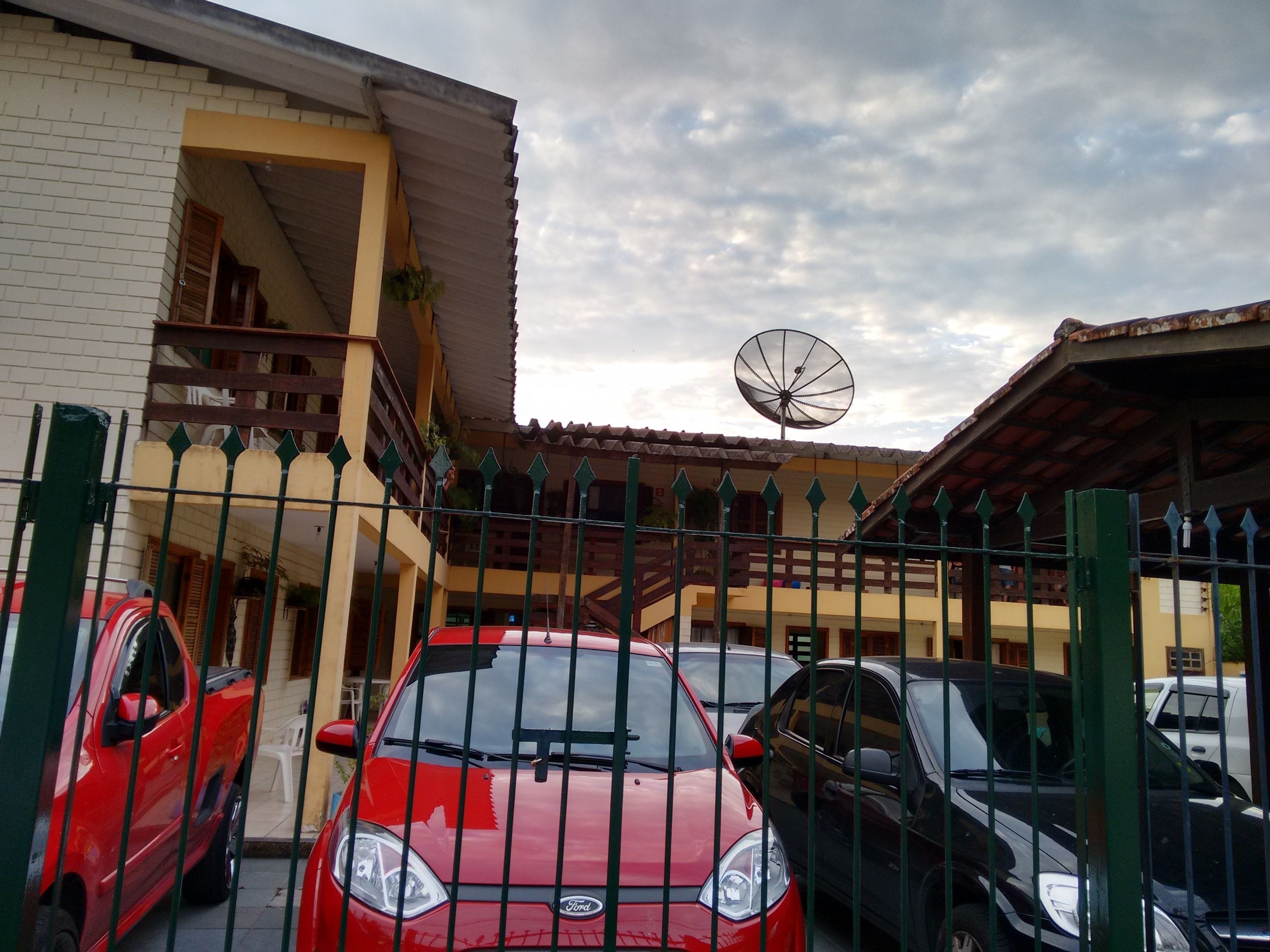 Uma área de estacionamento fechada por portão, com um carro vermelho e um carro preto estacionados em frente a um edifício com antena parabólica.
