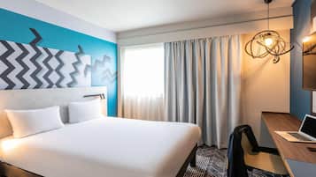 Quarto de hotel com uma cama grande, uma escrivaninha com um laptop, uma cadeira e um lustre moderno.