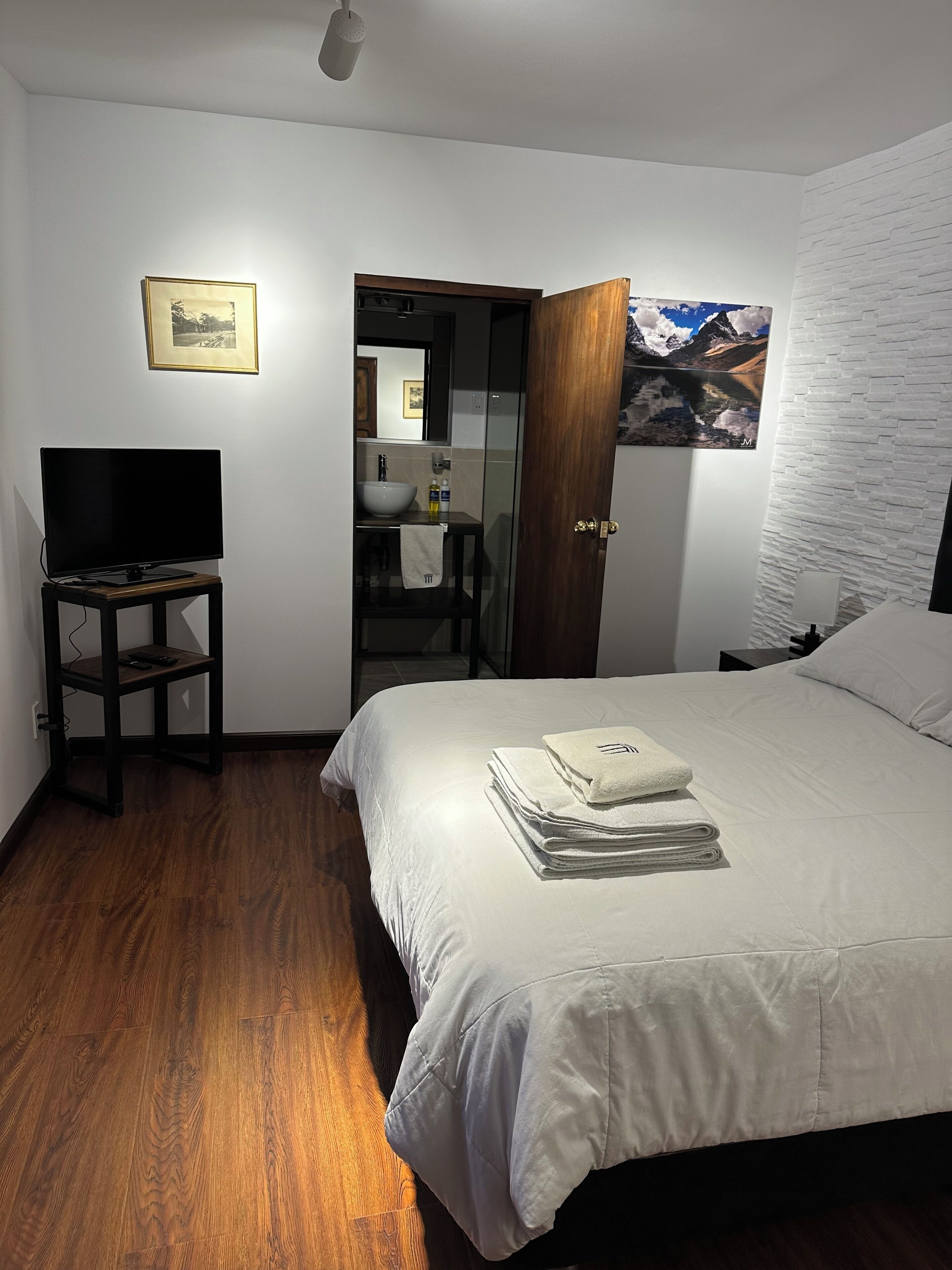Quarto de hotel com cama, televisão sobre um suporte de madeira, banheiro com espelho e um quadro na parede.