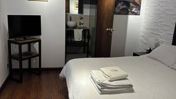 Quarto de hotel com cama, televisão sobre um suporte de madeira, banheiro com espelho e um quadro na parede.