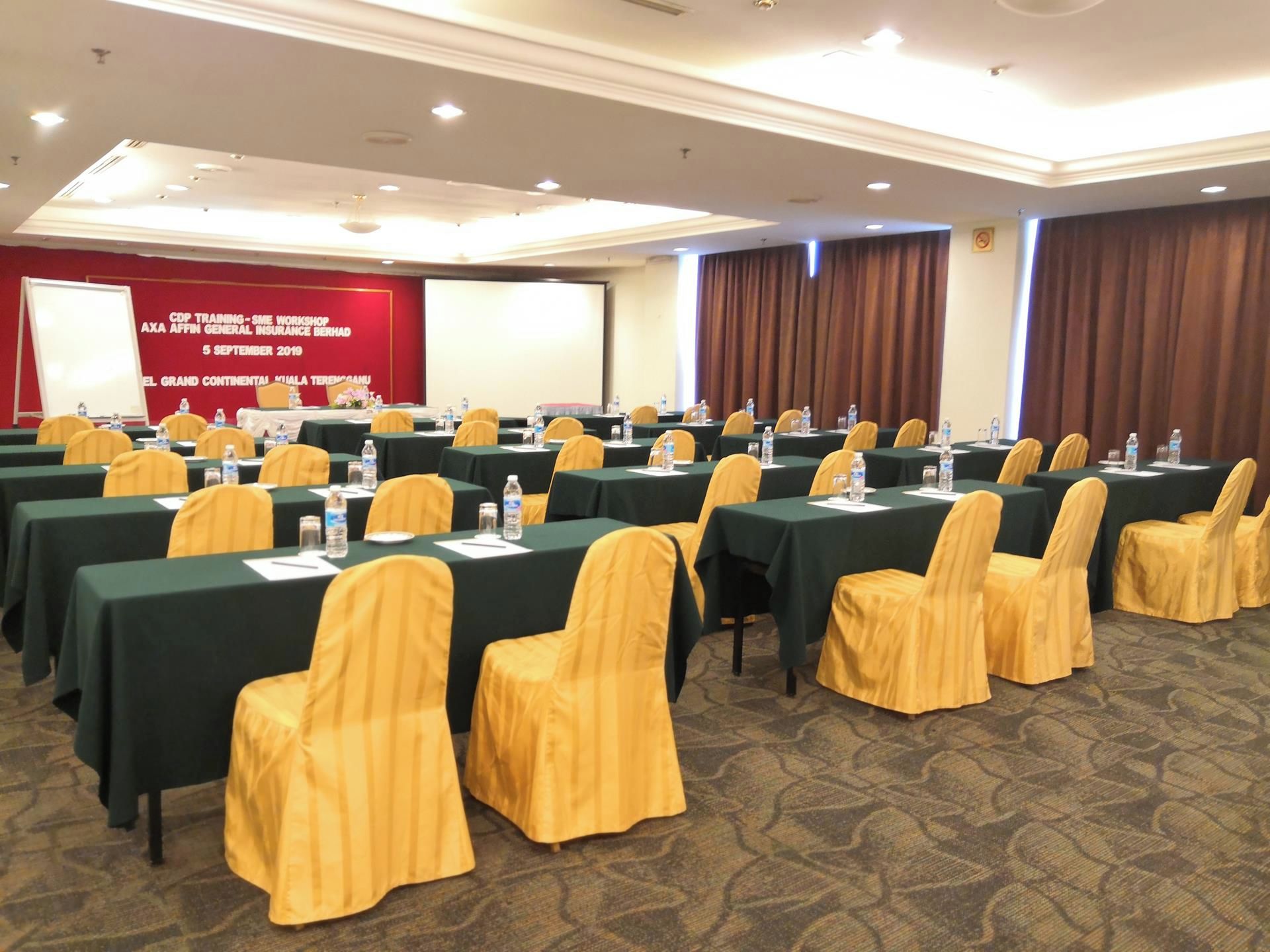 Sala de conferência com várias mesas e cadeiras, palco com tela de projeção e um banner com texto.