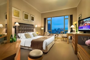 Quarto de hotel com uma cama grande, uma mesa de trabalho, televisão e vista para a cidade.