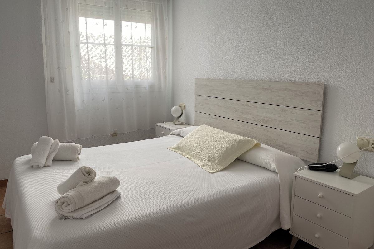 Uma cama bem arrumada com toalhas dobradas, uma luminária de cabeceira e uma mesa de cabeceira com carregador de celular.