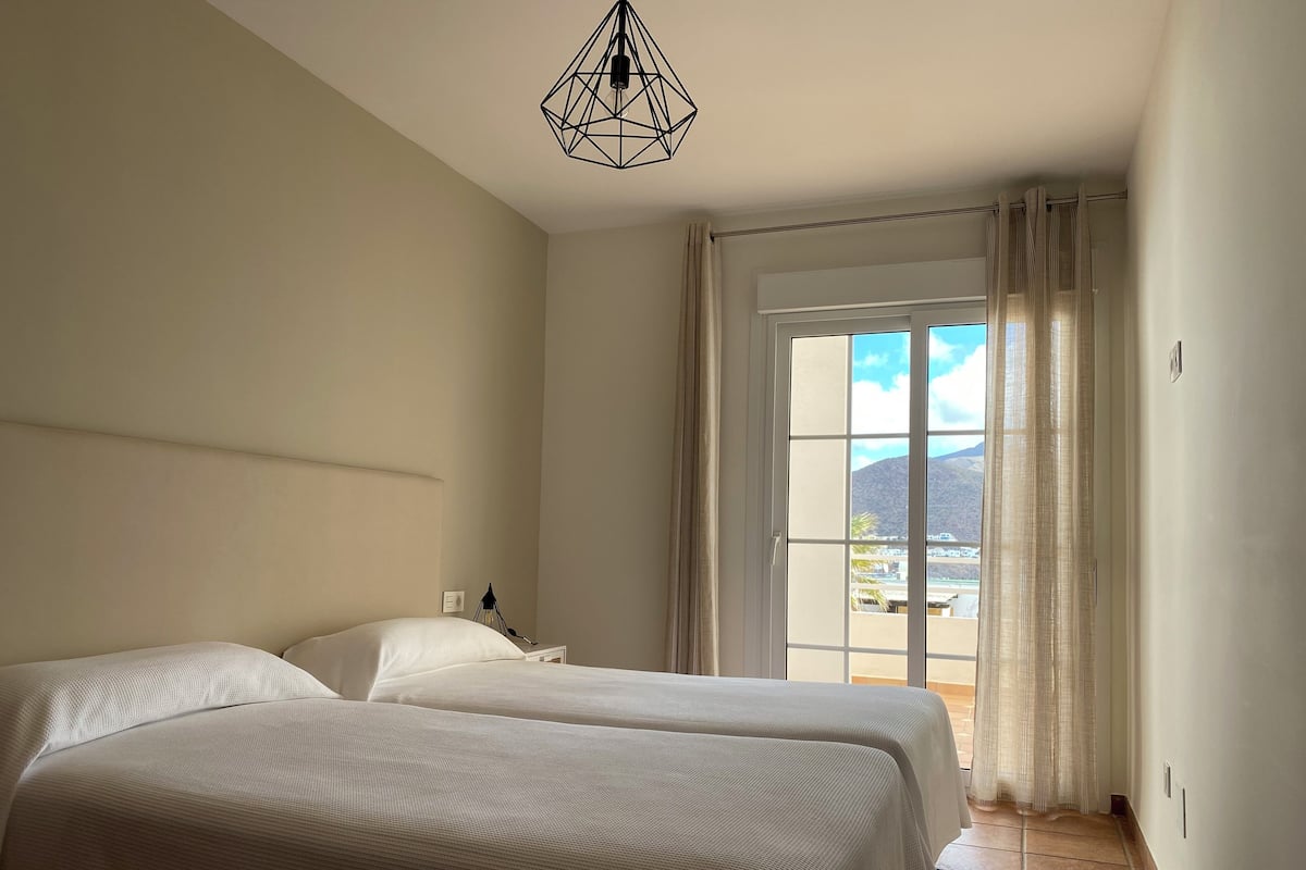 Quarto com duas camas, uma luminária de teto geométrica, uma janela com cortinas e vista para uma montanha.