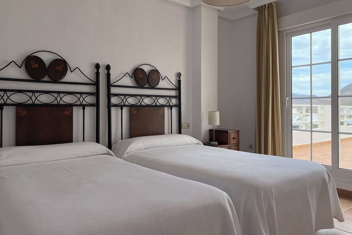 Quarto de hotel com duas camas, uma mesinha de cabeceira, uma luminária e uma janela grande com vista para a cidade.