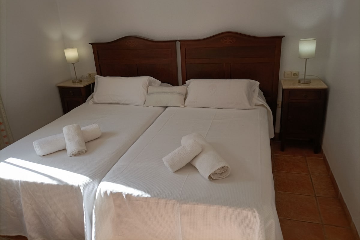 Cama de casal com duas almofadas e duas toalhas dobradas. Há duas mesas de cabeceira com abajures.