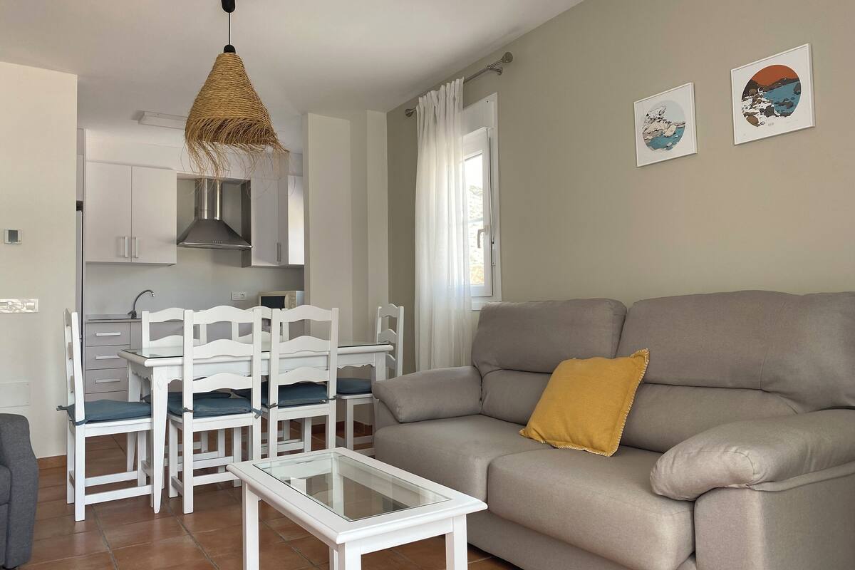 Uma sala moderna com um sofá cinza, uma mesa de centro de vidro e cadeiras brancas. Há uma área de cozinha com armários e um lustre.