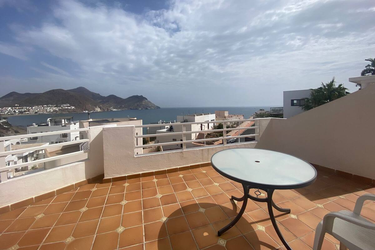 Terraço com mesa e cadeiras redondas, oferecendo vista para a costa, com edifícios e montanhas.