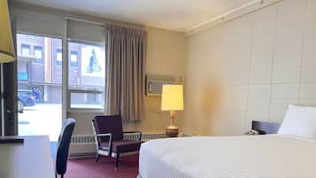 Quarto de hotel com cama, escrivaninha, cadeira, abajur e janela com cortinas.
