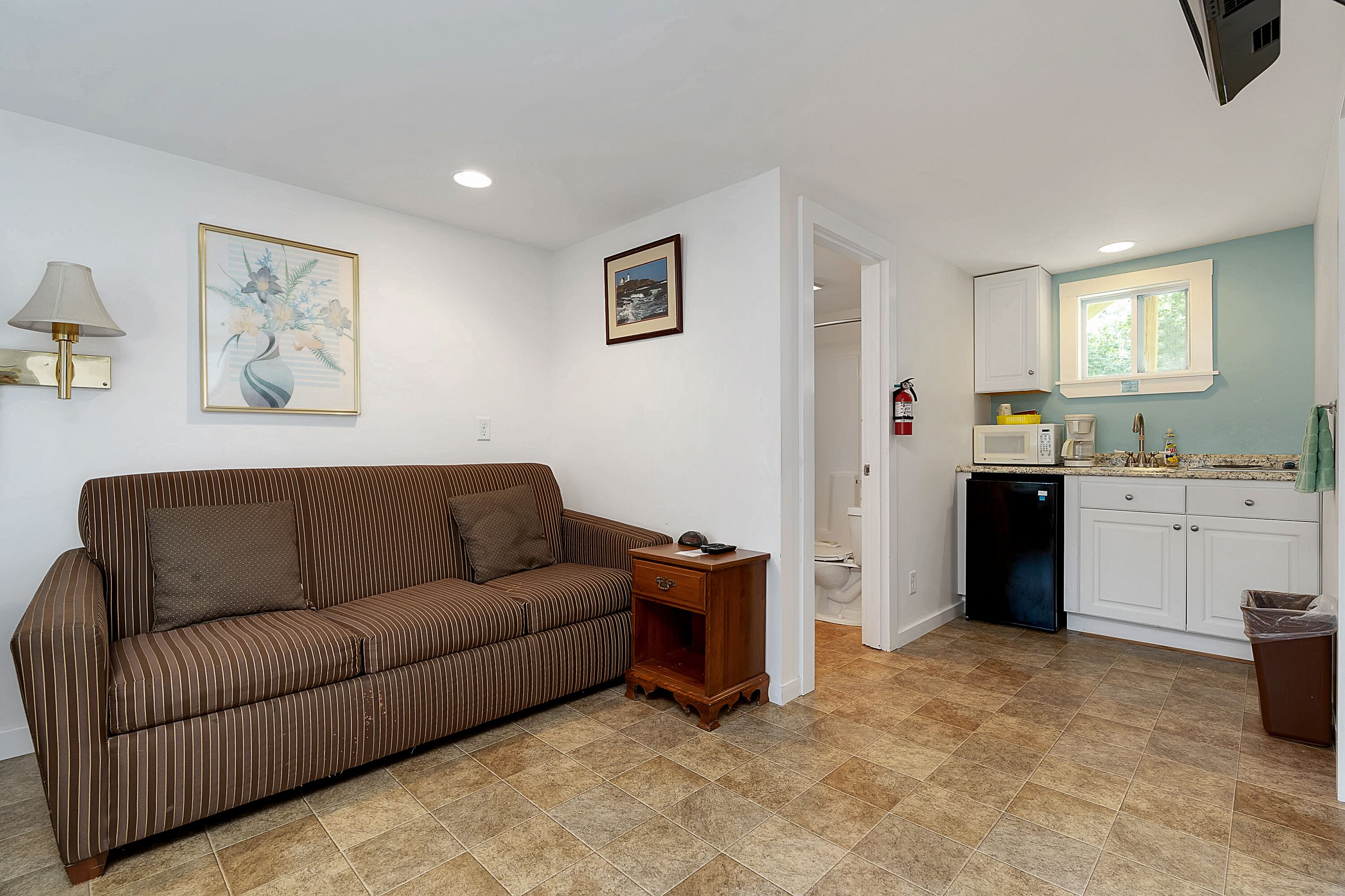Uma sala de estar com sofá listrado, uma pequena mesa lateral de madeira e um espaço de cozinha com armários brancos e micro-ondas.