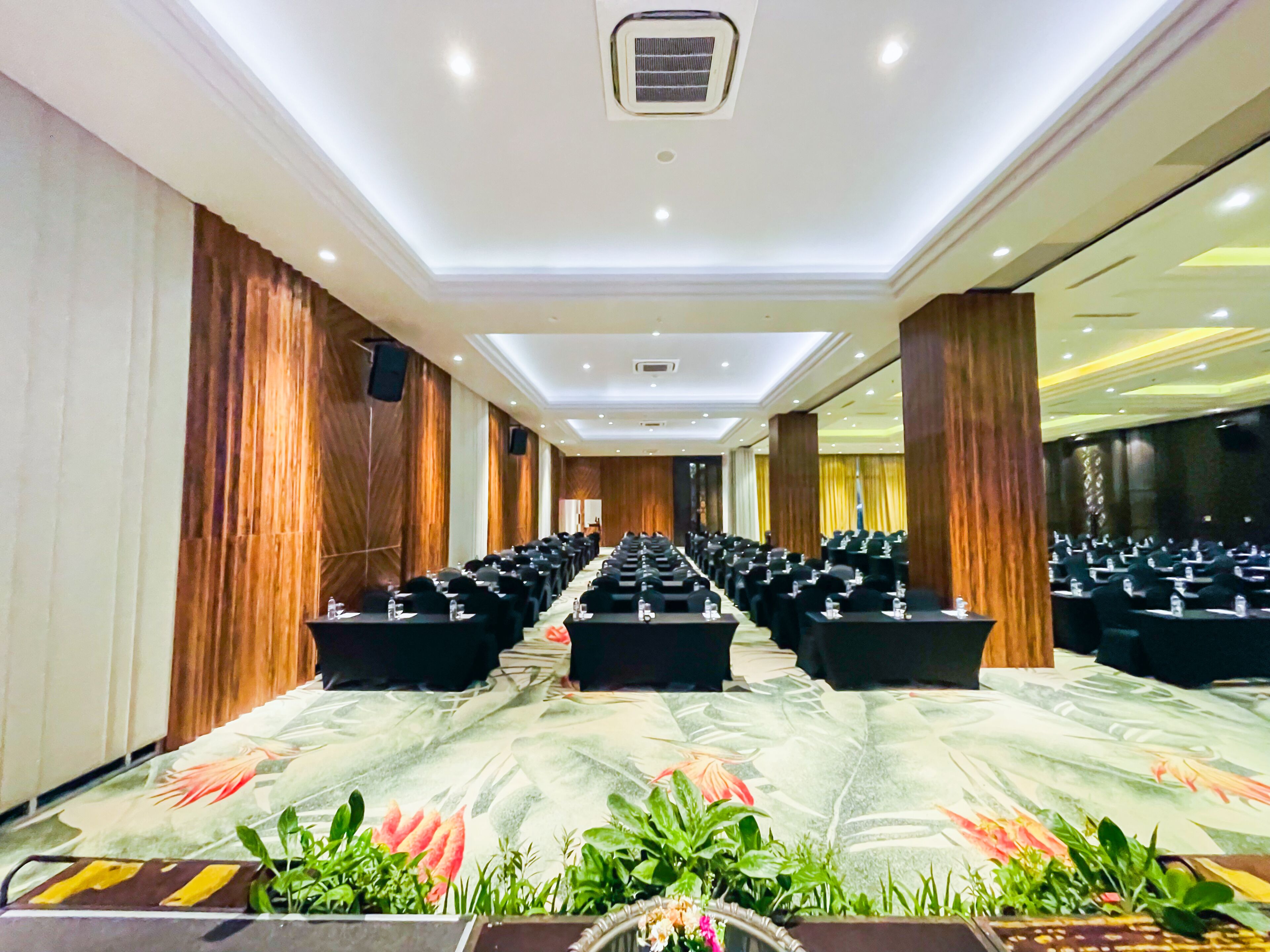 Uma sala de conferências com fileiras de mesas e cadeiras, carpete floral e painéis de madeira nas paredes.
