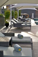 Área de estar ao ar livre iluminada pelo sol, com mobiliário de vime, piscina e plantas em vasos.