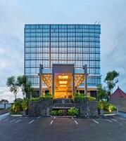 Um edifício moderno com fachada de vidro, entrada central com galeria coberta e estacionamento em frente.