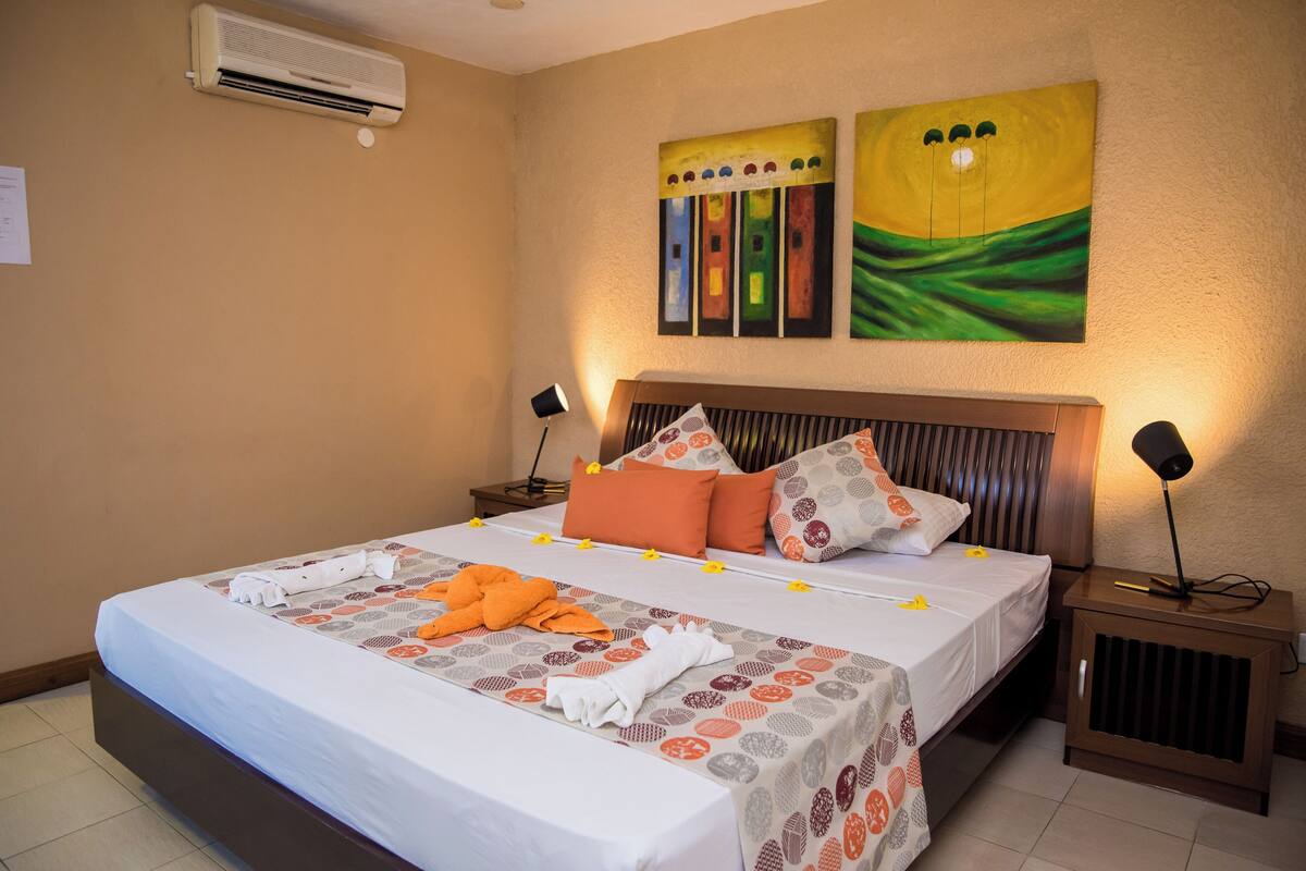 Quarto de hotel com uma cama, duas mesas de cabeceira, um ar condicionado e duas pinturas na parede.