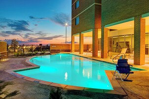 Uma piscina com borda curva, rodeada por um edifício de tijolo com amplas janelas e área de estar ao ar livre.