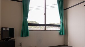 Quarto com tatames, uma televisão sobre um suporte e vista para uma colina através de uma janela com cortinas verde-água.