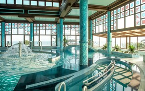Piscina interna com cascata, rodeada por amplas janelas com padrões geométricos e vista para o oceano.