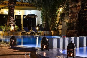 Uma área de piscina à noite, com espreguiçadeiras, um espaço de estar coberto e lanternas decorativas.