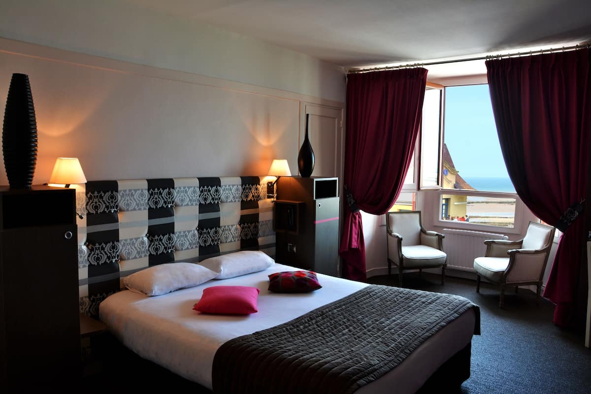 Quarto de hotel com uma cama grande, duas luminárias de cabeceira, uma janela com cortinas, uma cadeira e uma pequena mesa.