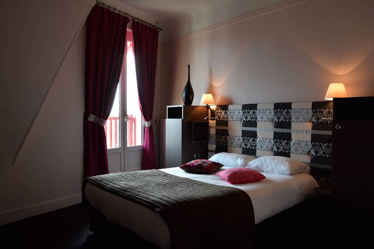 Quarto de hotel com cama, mesa de cabeceira com duas luminárias, janela com cortinas vermelhas e cabeceira estampada.